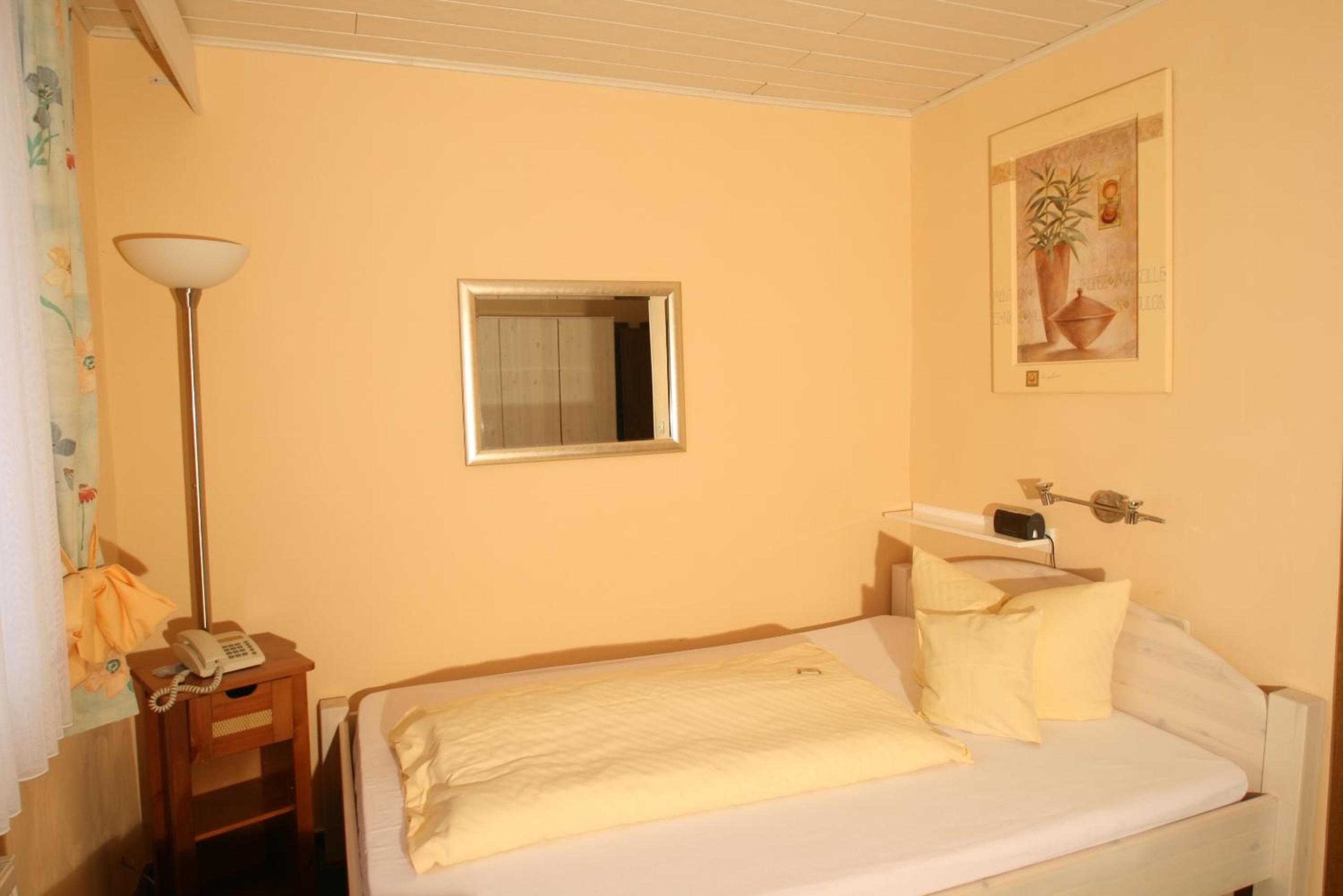Quarto de hotel com cama, mesa de cabeceira com abajur, telefone, pequena janela e um quadro na parede.