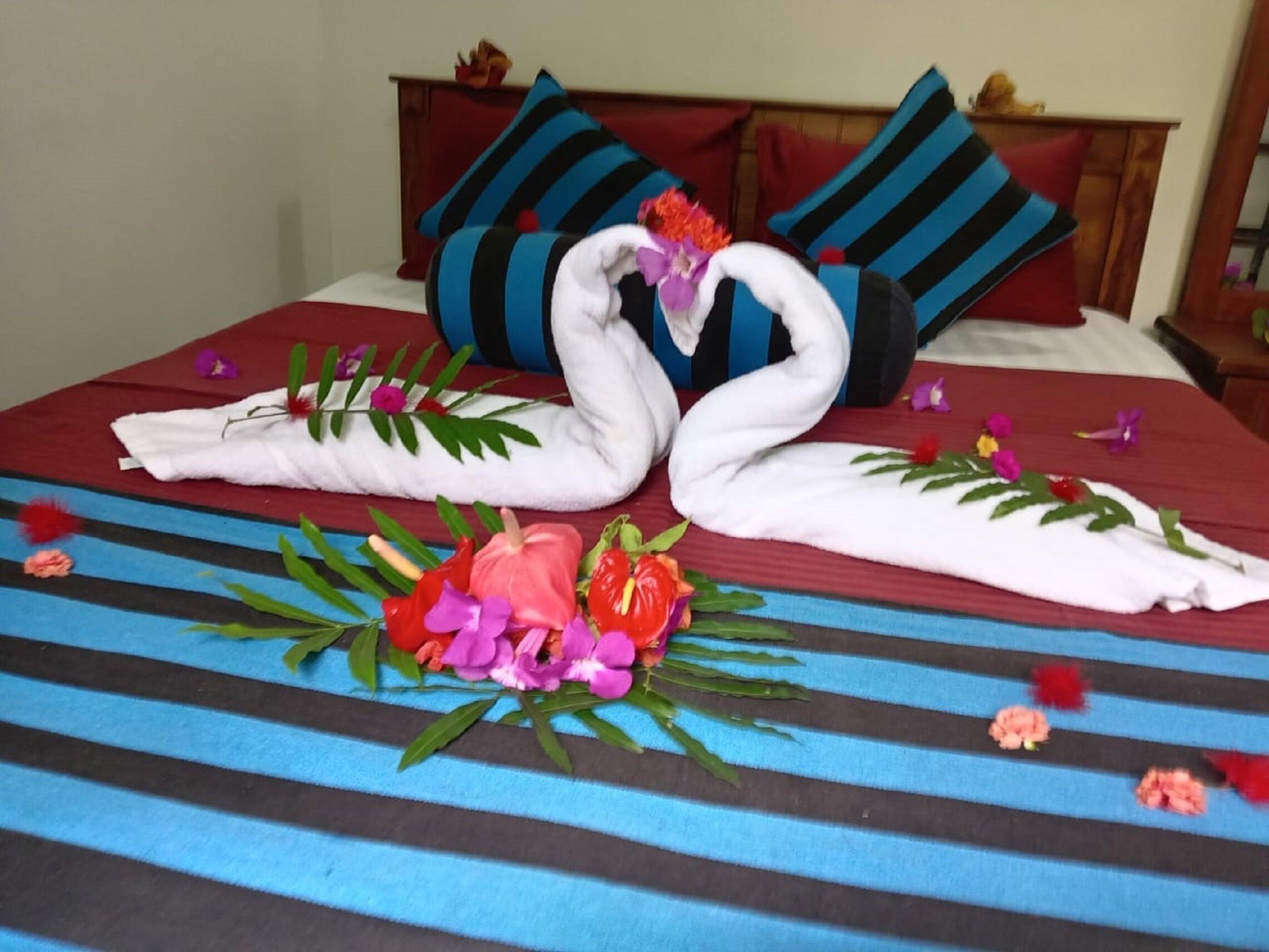 Cama com colcha listrada em vermelho e azul, duas toalhas dobradas em formato de cisne e um arranjo floral sobre a cama.