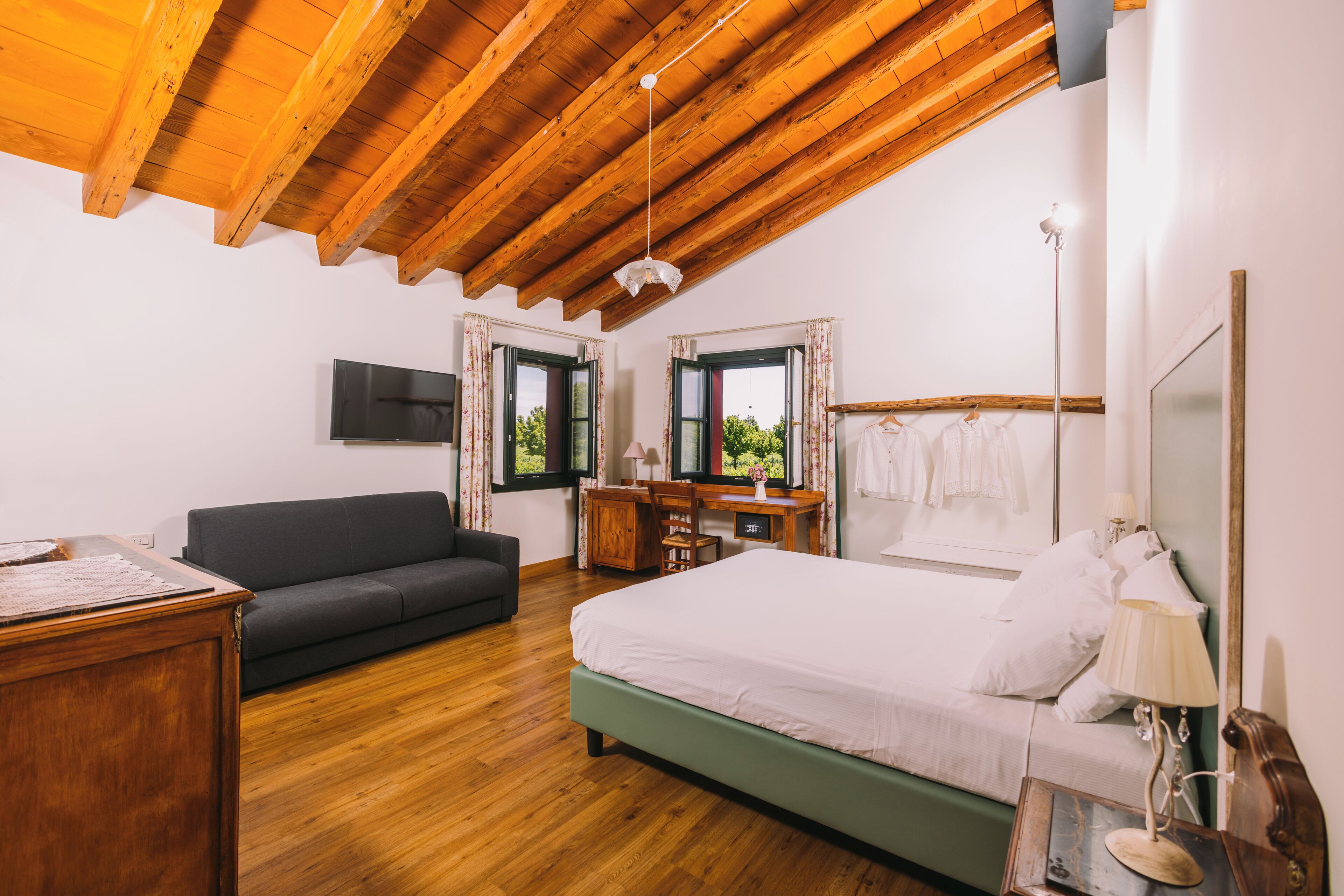 Quarto com teto de madeira, uma cama, escrivaninha, cadeira, sofá, televisão e janela com cortinas.