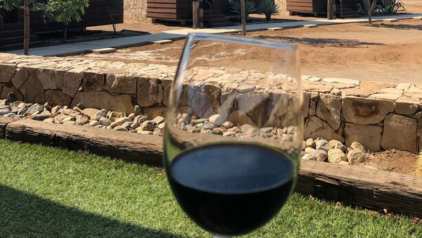 Um copo de vinho tinto em primeiro plano, com vista para um edifício moderno e uma parede de pedra ao fundo.