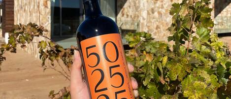 Uma mão segura uma garrafa de vinho com etiqueta, em frente a um edifício moderno com elementos de pedra e vidro.
