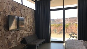 Quarto com parede de pedra, TV de tela plana, sofá, cama e vista para um vinhedo por grandes janelas.