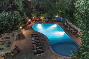 Piscina com espreguiçadeiras e guarda-sóis, rodeada por árvores e pavimento de pedra.