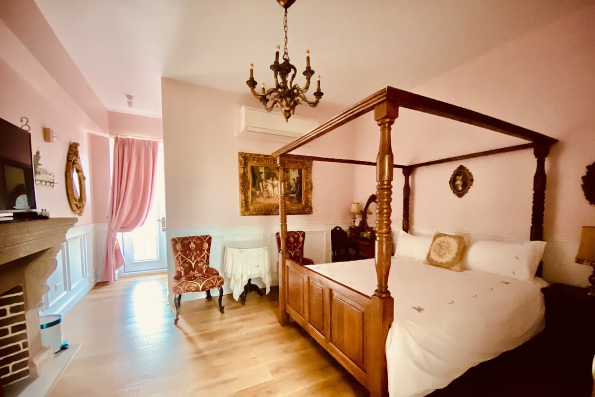 Uma cama com dossel e estrutura de madeira, um candelabro, uma lareira e uma poltrona com padrão floral.