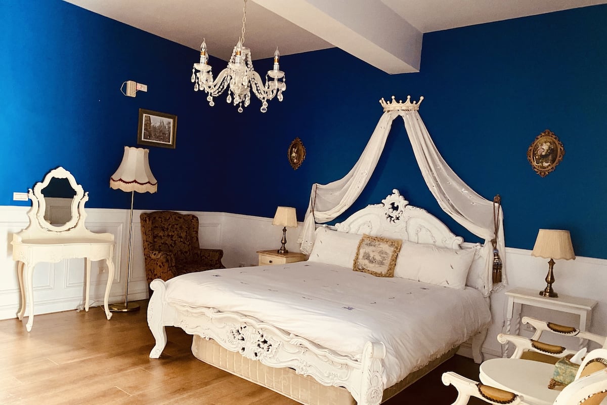 Quarto com cama de dossel, lustre, espelho, cadeira e mesa de cabeceira com abajur.