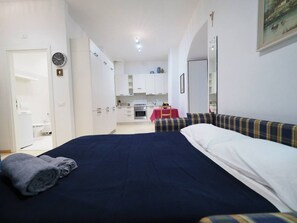 Quarto de hotel com uma cama, uma mesa vermelha e uma cozinha compacta ao fundo.