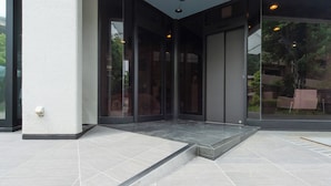 Entrada de um edifício moderno com porta de vidro, uma pequena área de estar ao ar livre e piso ladrilhado.