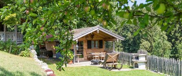 Uma cabana de madeira com telhado inclinado, um alpendre com mobiliário de exterior e uma cerca de madeira delimitando a propriedade.