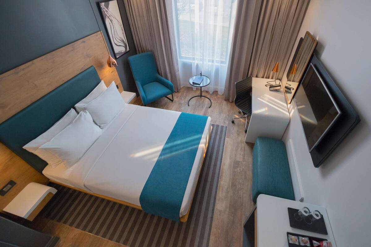 Quarto de hotel com cama, escrivaninha, cadeira, televisão e mesa pequena.