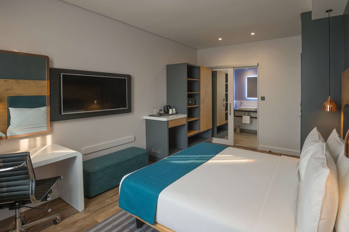 Quarto de hotel moderno com uma cama grande, escrivaninha, cadeira, guarda-roupa e banheiro visível através de uma porta aberta.