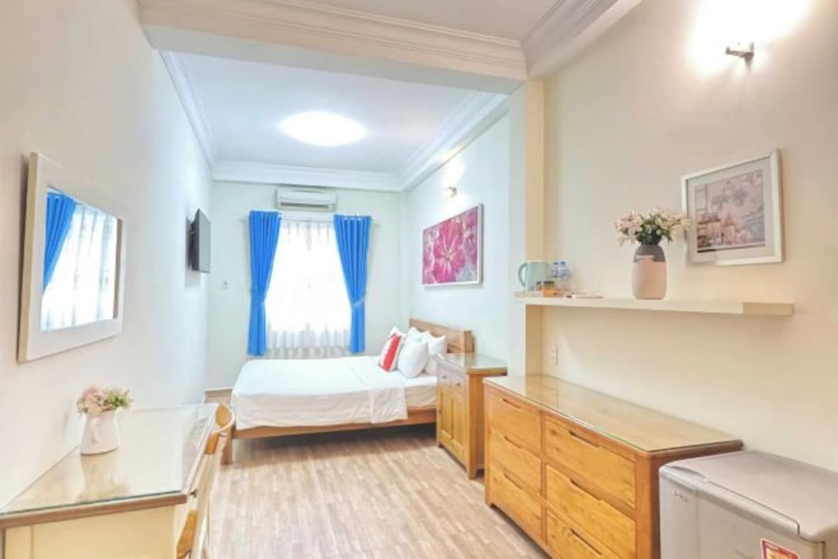 Quarto de hotel com cama, escrivaninha, cômoda, televisão e prateleira fixada na parede com um vaso de flores.