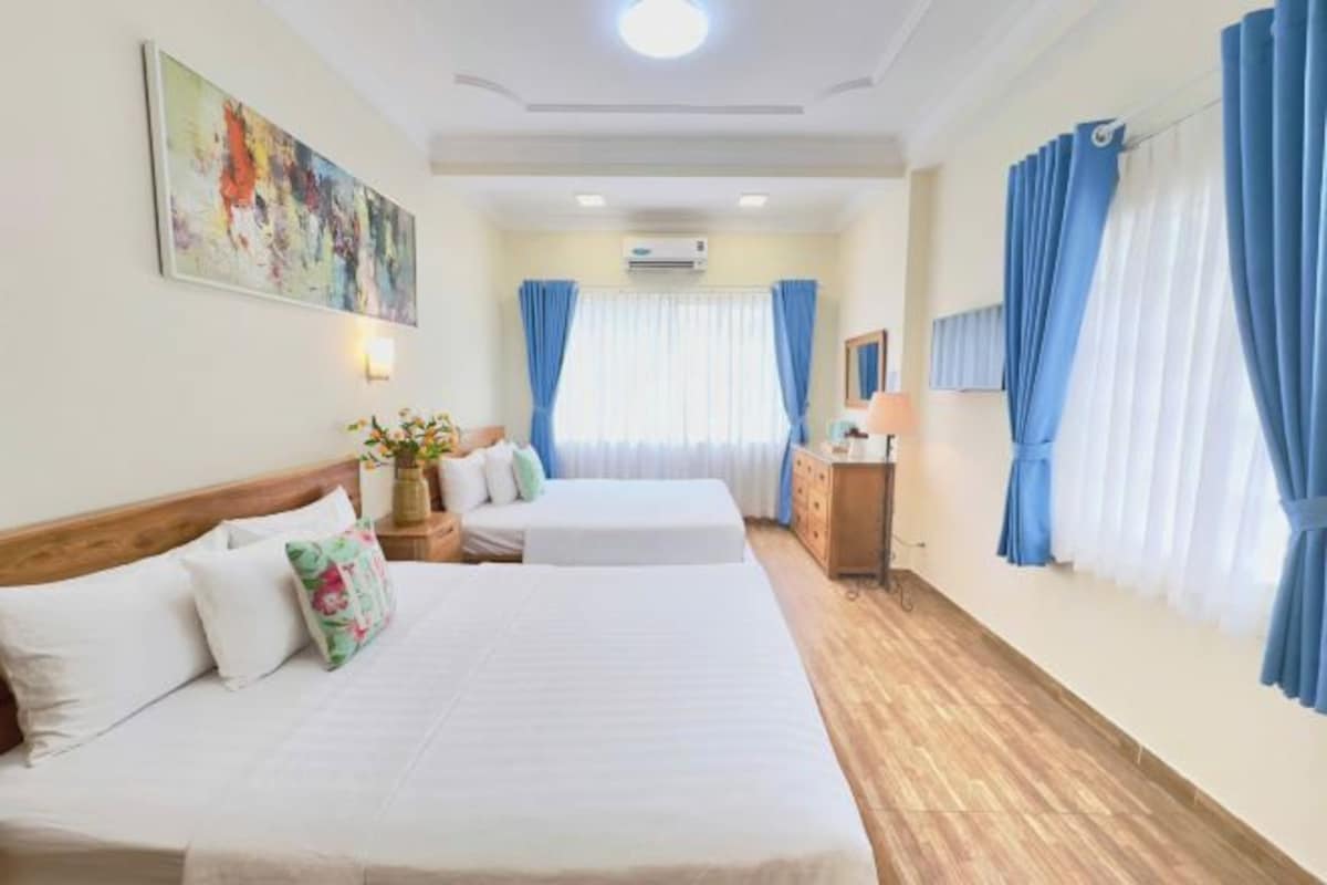 Quarto de hotel com duas camas, cabeceira de madeira, mesa de cabeceira com abajur, cômoda e um quadro na parede.