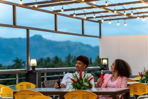 Um casal jantando em uma mesa ao ar livre, vista para as montanhas, cadeiras amarelas e luzes de festa.