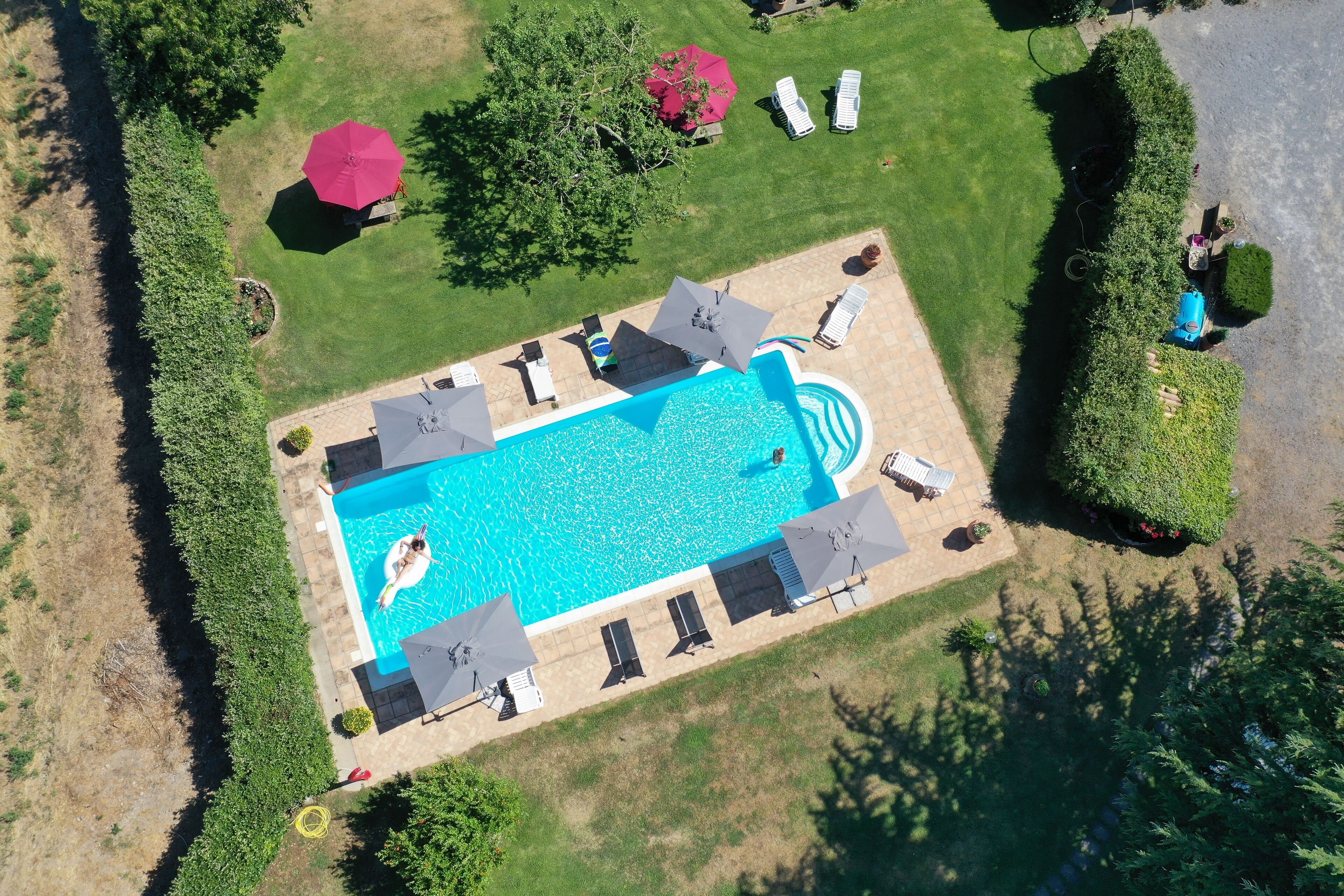 Vista aérea de uma piscina retangular com deck ladrilhado, cercada por sebes bem aparadas e algumas cadeiras de descanso.