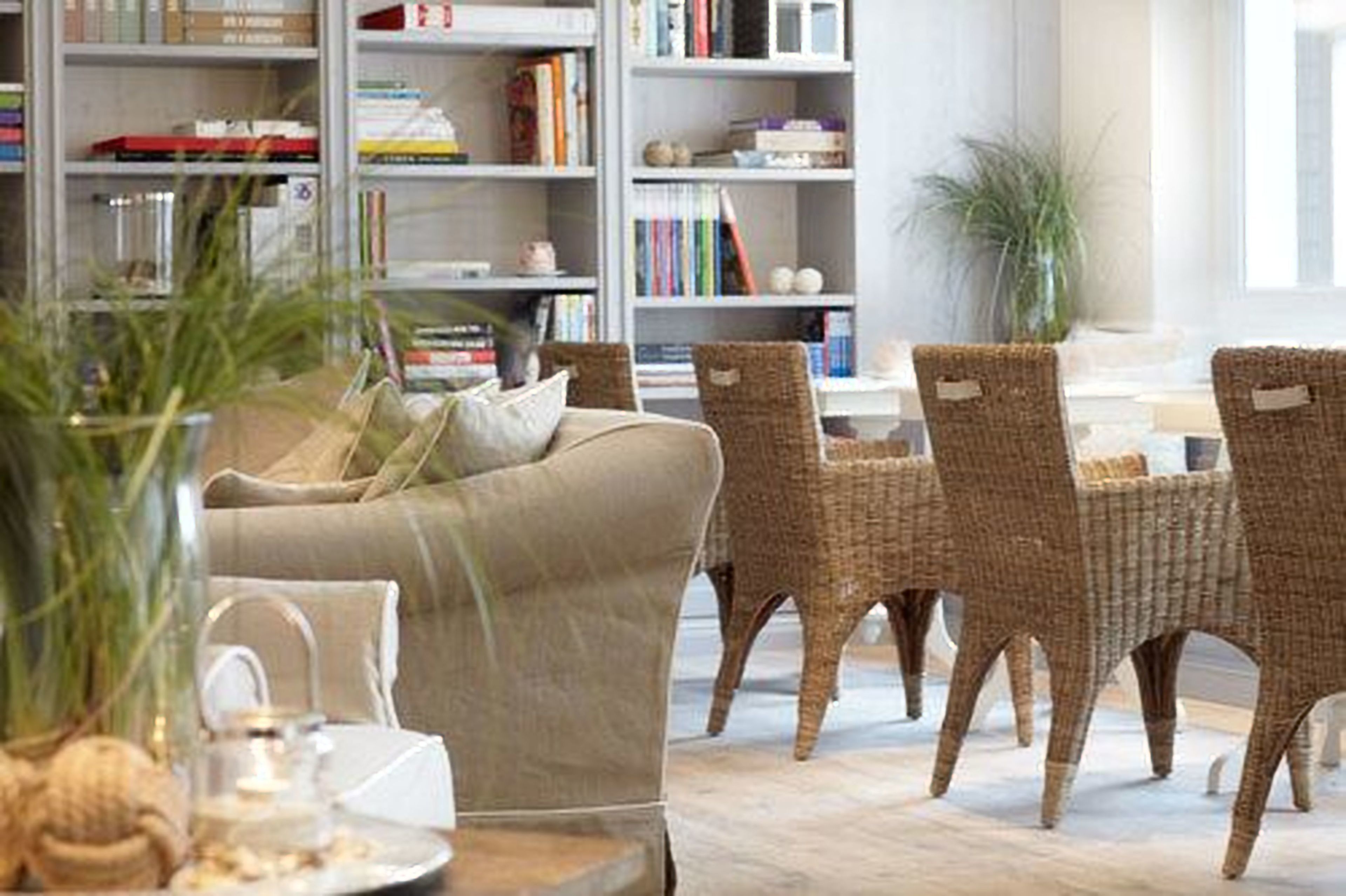 Uma sala de estar com um sofá bege, cadeiras de vime, uma mesa de centro e uma estante cheia de livros.