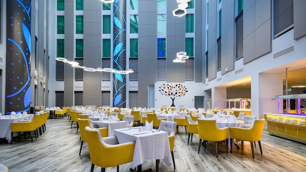 Uma ampla área de refeições com cadeiras amarelas e mesas brancas, um mural decorativo com tema de árvore no centro, e luminárias modernas.