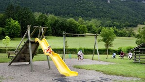 Um parquinho com um escorrega amarelo e balanços, numa área verde com árvores.