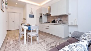 Cozinha moderna com armários brancos, forno embutido e fogão. Possui uma área de refeições com mesa e cadeiras brancas, e um tapete estampado no chão.