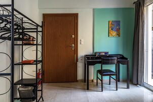 Quarto com porta de madeira, uma escrivaninha com cadeira, estante e pequeno armário branco.