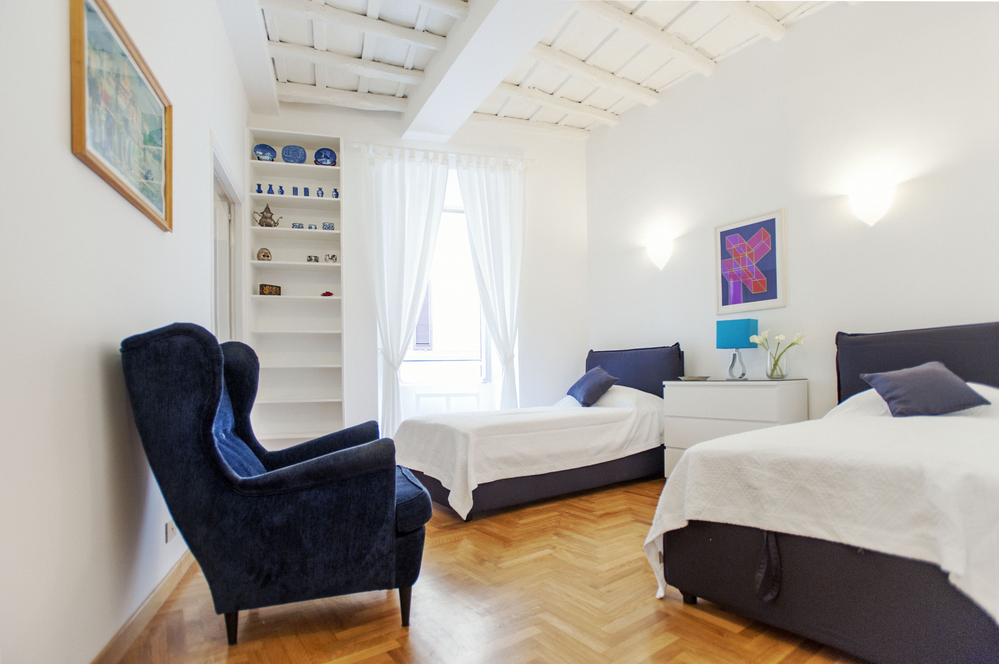 Quarto com duas camas, uma cadeira, mesa de cabeceira com abajur, prateleira com objetos decorativos e janela com cortinas.