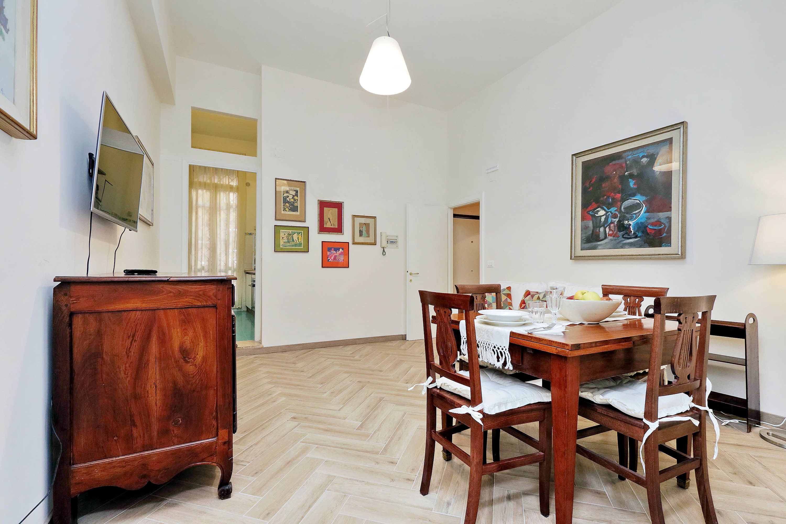 Uma sala de jantar com mesa de madeira, cadeiras e um armário. Há quadros na parede e uma pintura sobre a mesa.