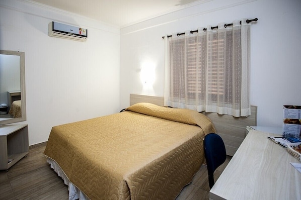 Quarto de hotel com cama, escrivaninha, cadeira, espelho e janela com cortinas.