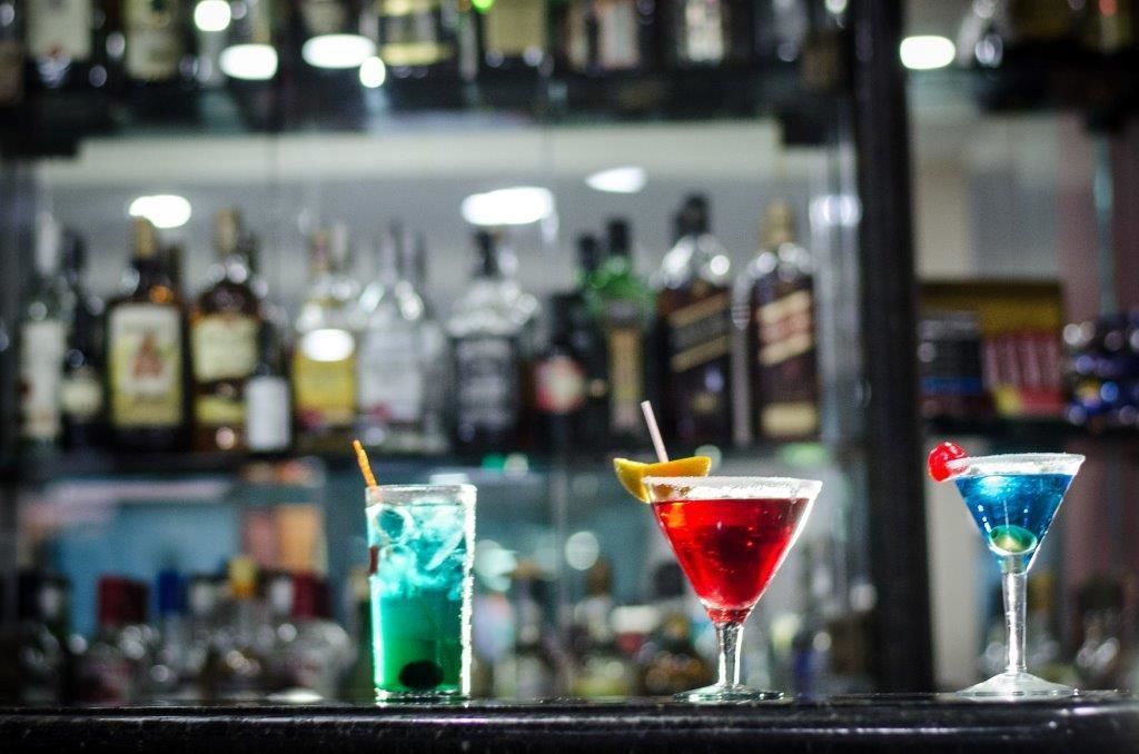 Três cocktails sobre um balcão de bar, com um fundo desfocado de diversas garrafas.