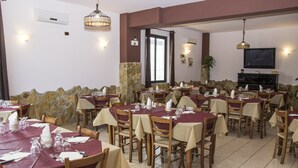 Restaurante com várias mesas preparadas para refeições, uma televisão presa à parede e uma parede de pedra ao fundo.