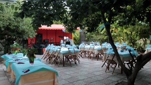 Uma área de refeições ao ar livre com mesas dispostas sob árvores, um palco com equipamentos musicais e uma tenda vermelha ao fundo.