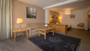 Quarto de hotel com sofá, mesa de centro de madeira, pequena mesa com luminária, cama, cadeira e área de cozinha.
