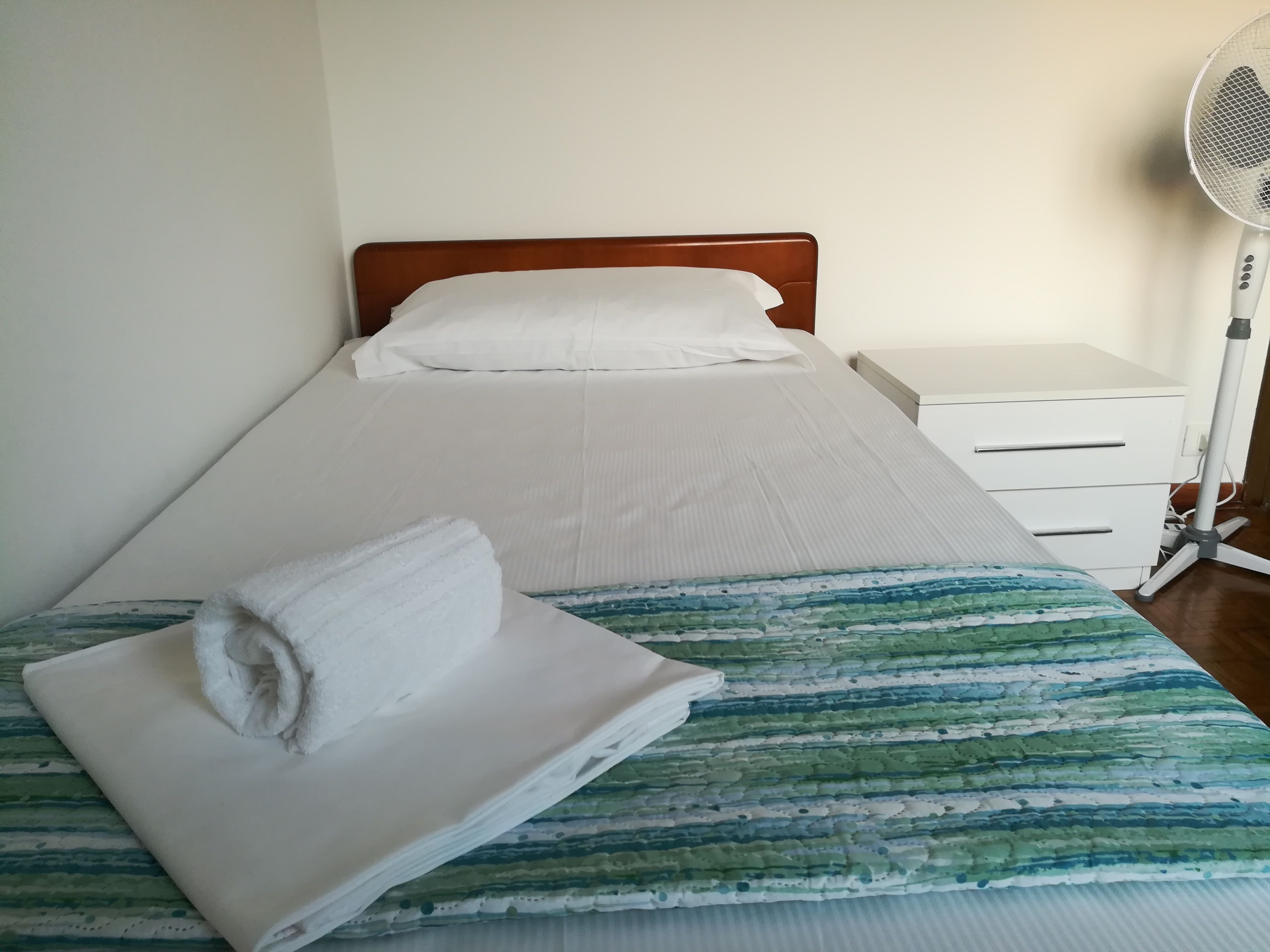 Cama bem arrumada com lençóis brancos e uma toalha dobrada, mesa de cabeceira com abajur branco e colcha listrada.