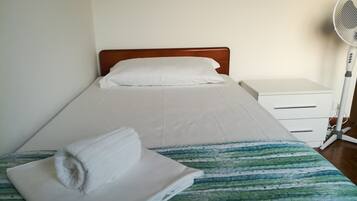 Cama bem arrumada com lençóis brancos e uma toalha dobrada, mesa de cabeceira com abajur branco e colcha listrada.