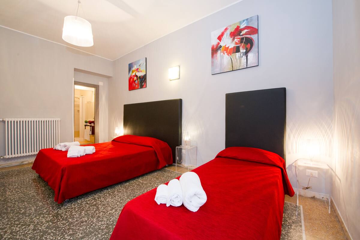 Quarto de hotel com duas camas, roupa de cama vermelha, radiador branco e obras de arte nas paredes.