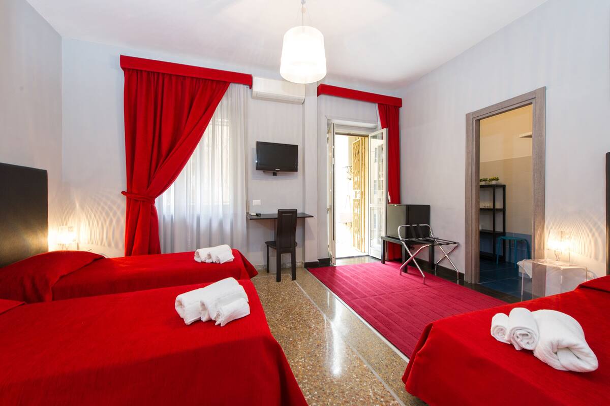 Quarto de hotel com duas camas, cortinas vermelhas, uma TV, uma escrivaninha e uma cadeira.