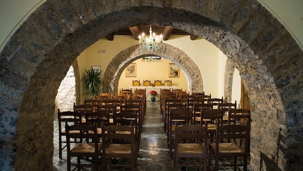 Um corredor de pedra arqueado com bancos de madeira dispostos em fileiras, conduzindo a uma área central com altar e um lustre.