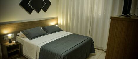 Uma cama bem arrumada com cabeceira, uma mesa de cabeceira com luminária e uma televisão sobre um suporte.