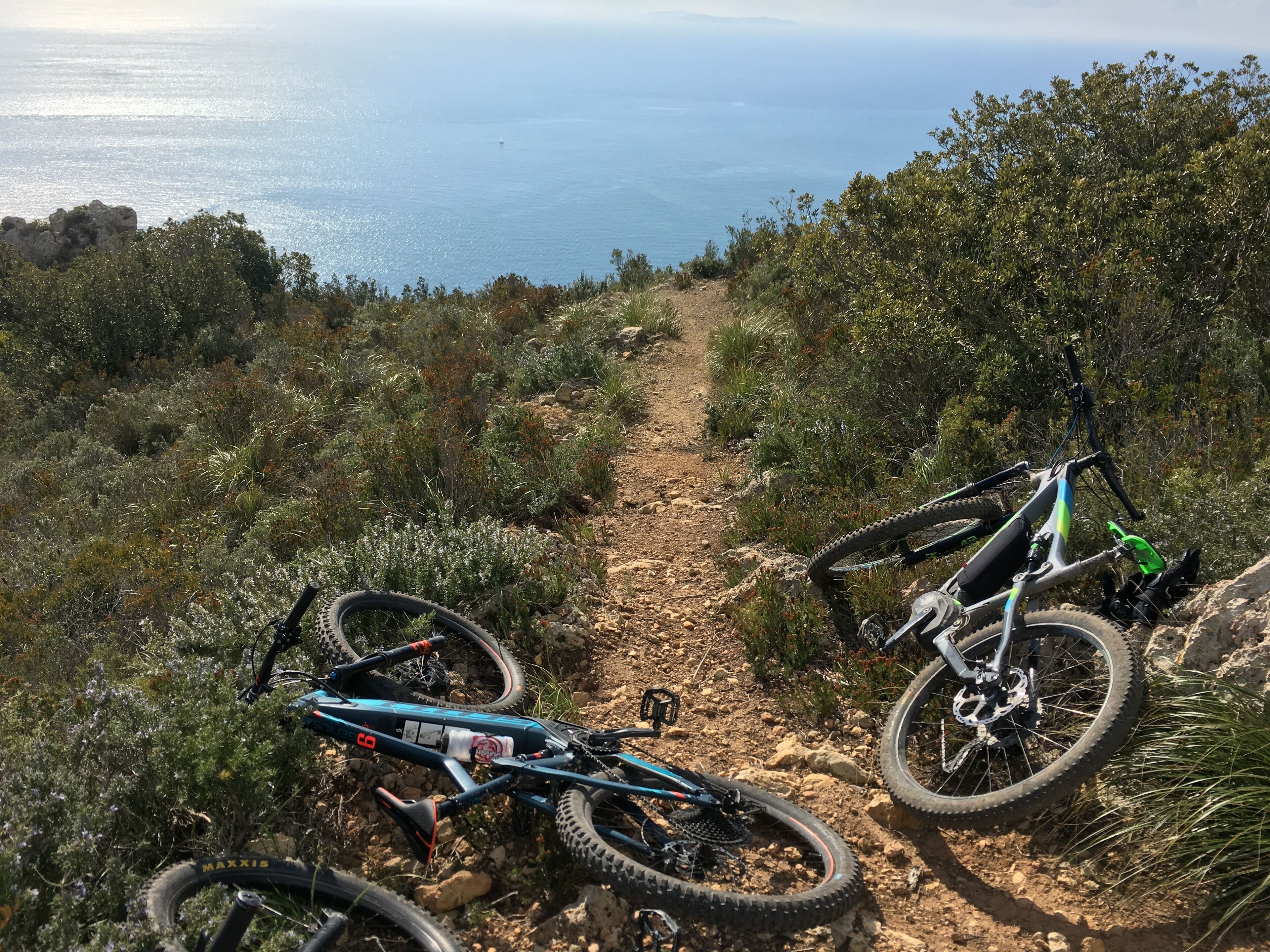 Trilha de bicicleta de montanha com duas bicicletas, em meio a terreno rochoso e vegetação, com vista para a costa.