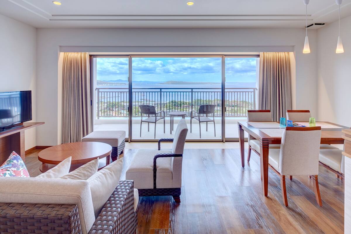 Quarto de hotel moderno com varanda, área de refeições e espaço de estar com televisão e sofá.