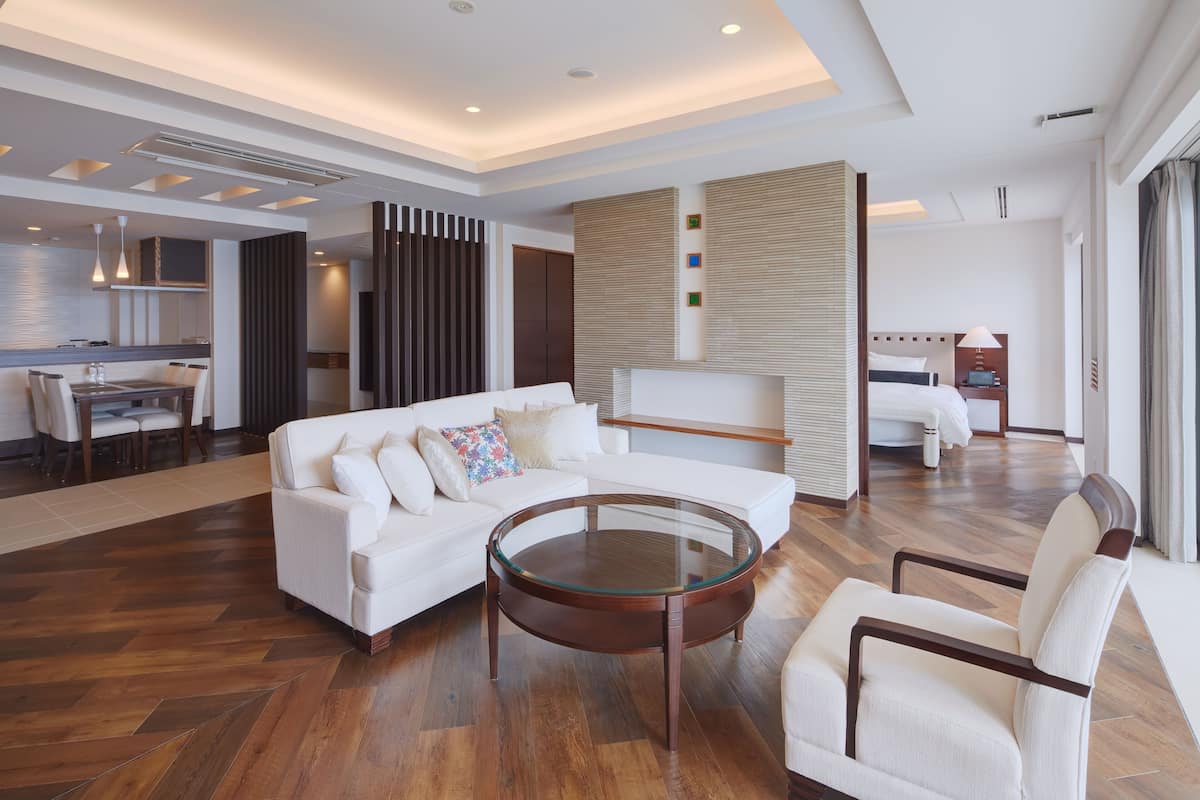 Quarto de hotel moderno com sofá branco, mesa de centro de vidro e piso de madeira.