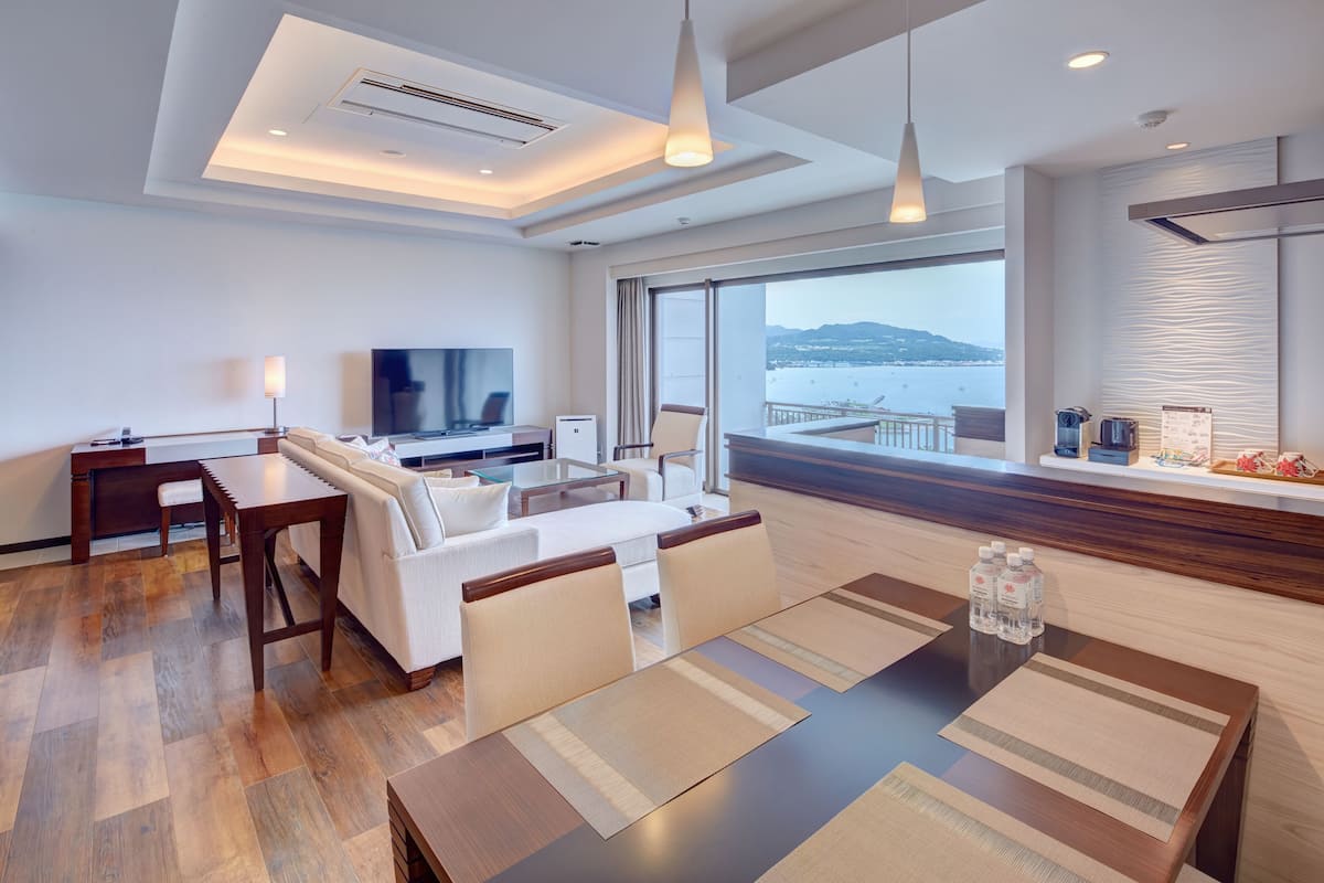 Quarto de hotel moderno com área de refeições, sofá, televisão e uma grande janela com vista para o mar.