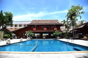 Piscina com deck ladrilhado, cercada por palmeiras e um edifício com telhado telhado.