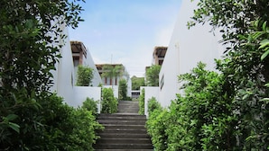 Escadaria levando a um edifício com paredes brancas e telhado de palha, cercado por vegetação exuberante.