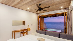 Quarto com cama, escrivaninha, cadeira, ventilador de teto e vista para o mar por uma porta aberta.