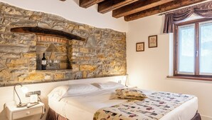 Um quarto com parede de pedra, cama com manta estampada, mesa de cabeceira com abajur e janela com cortinas.