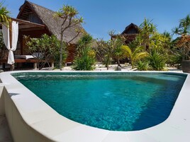 Uma piscina límpida com borda curva, rodeada por vegetação tropical e edifícios com telhados de palha.