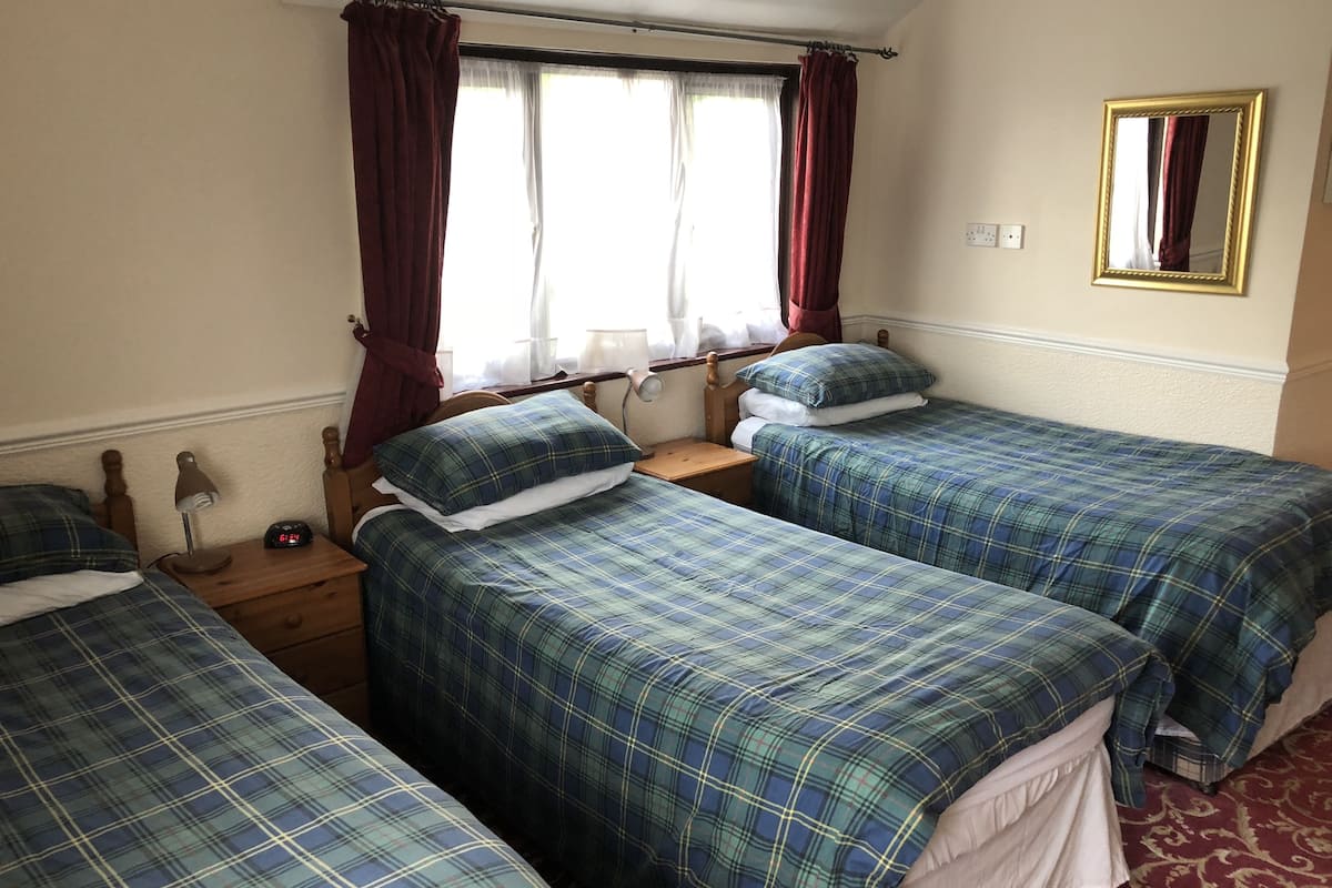 Quarto com duas camas de solteiro, mesa de cabeceira com luminária, relógio e janela com cortinas.