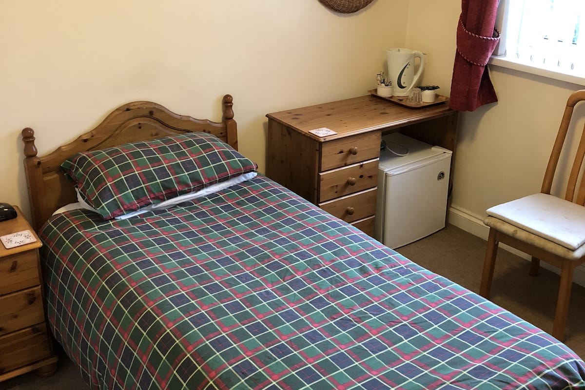 Quarto com cama, escrivaninha, cadeira, pequeno frigorífico, espelho e janela com cortinas.