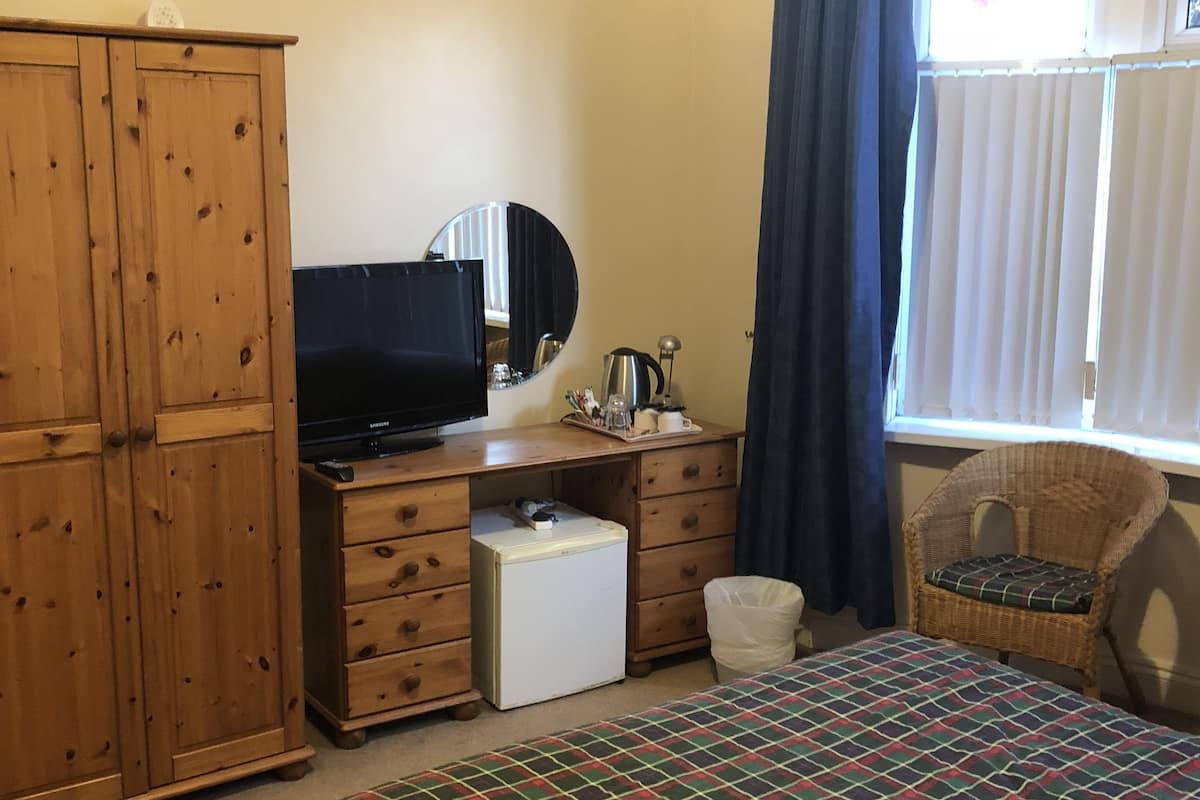 Quarto com cama, guarda-roupa de madeira, escrivaninha com computador, cadeira e janela com cortinas.