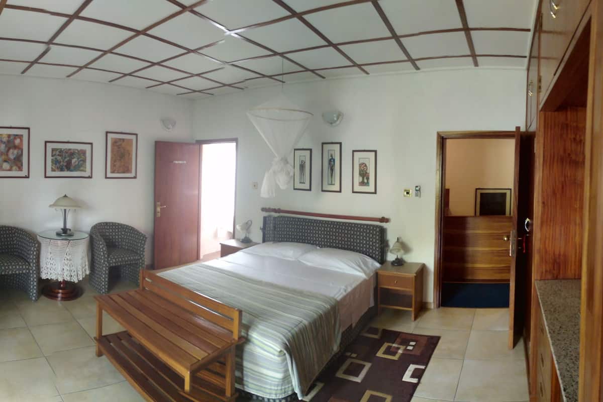 Quarto com cama, mesas de cabeceira, um banquinho, uma cadeira, guarda-roupa e uma porta.