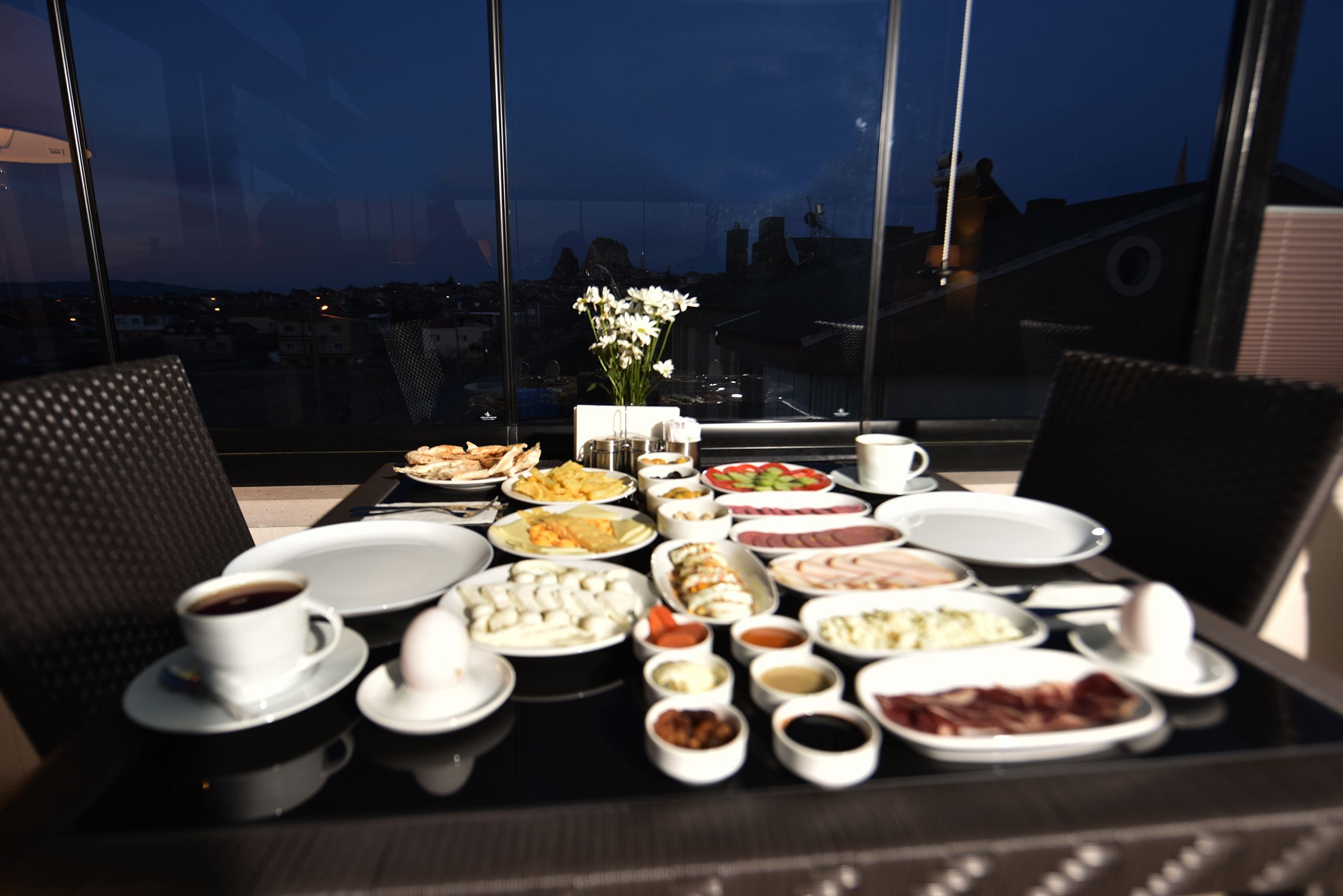 Mesa de jantar posta com diversos pratos e uma xícara de café, posicionada perto de uma janela com vista para a paisagem urbana ao entardecer.